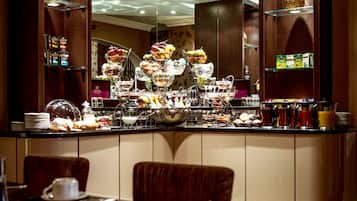 Um buffet de café da manhã com diversos alimentos e bebidas, uma área de estar com mesas e cadeiras, e um teto decorativo com iluminação embutida.