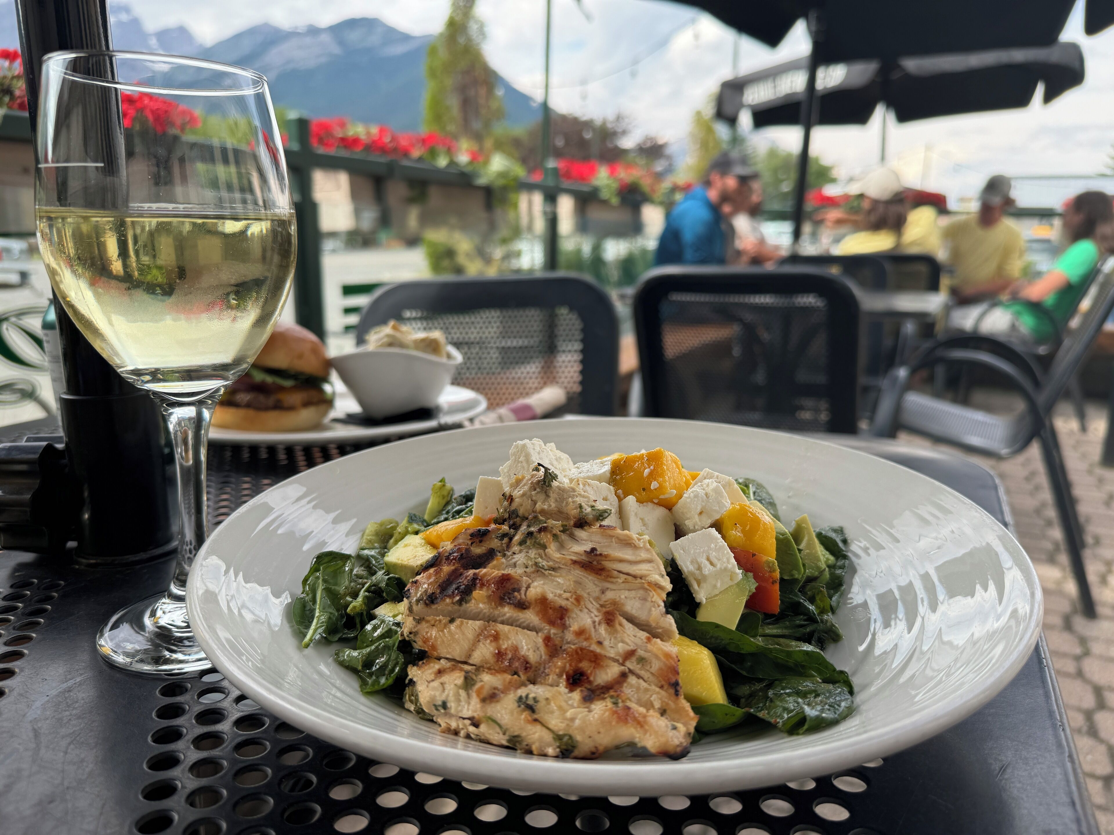 Uma refeição com frango grelhado e legumes num prato branco, um copo de vinho branco e uma mesa com padrão de grelha metálica.