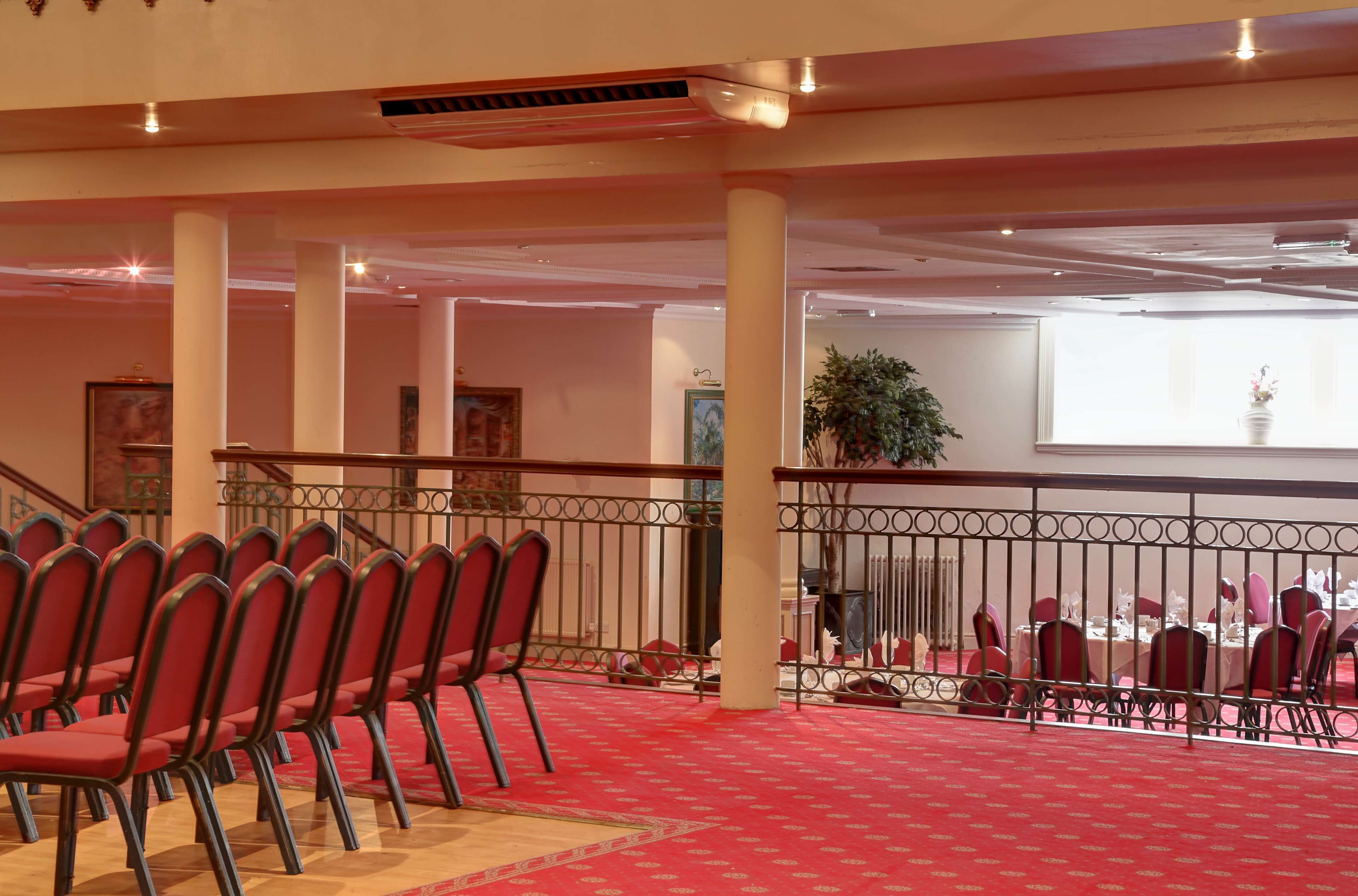 Uma sala de conferências com fileiras de cadeiras vermelhas, um tapete vermelho e uma varanda com mesas preparadas para refeições.