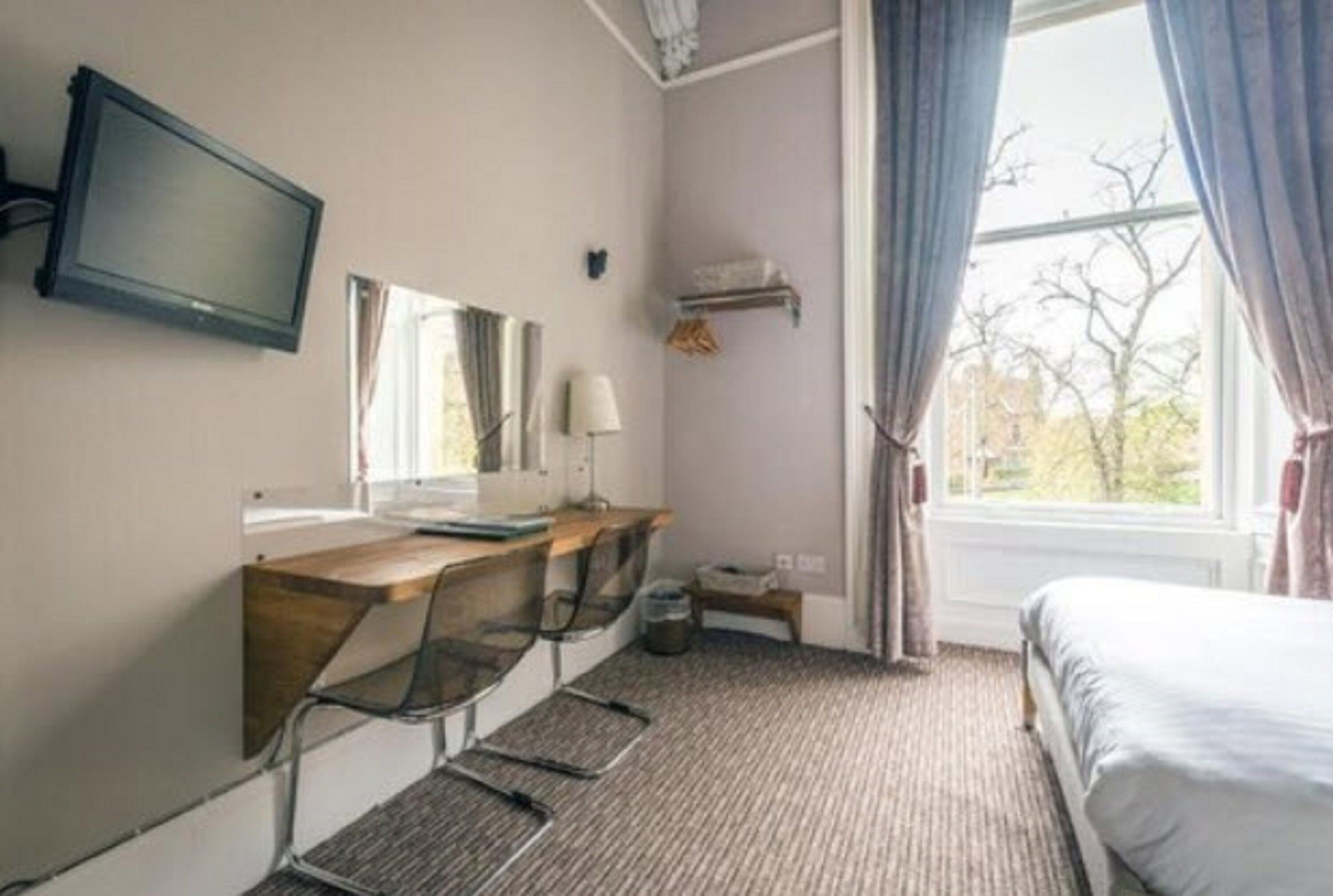 Quarto de hotel com cama, escrivaninha com cadeira, televisão na parede, janela com cortinas e pequena estante com objetos.