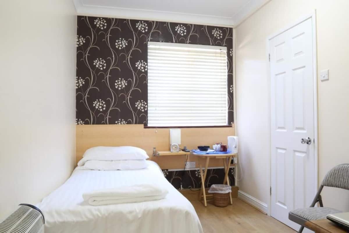 Um quarto pequeno e bem organizado, com uma cama, escrivaninha e cadeira, janela com persianas e porta.