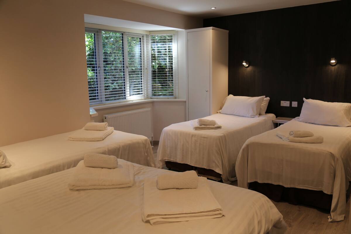 Quarto de hotel com três camas simples, cada uma com colcha branca e toalha dobrada. Há uma janela com persianas, um radiador sob a janela e uma parede escura com duas luzes de parede.