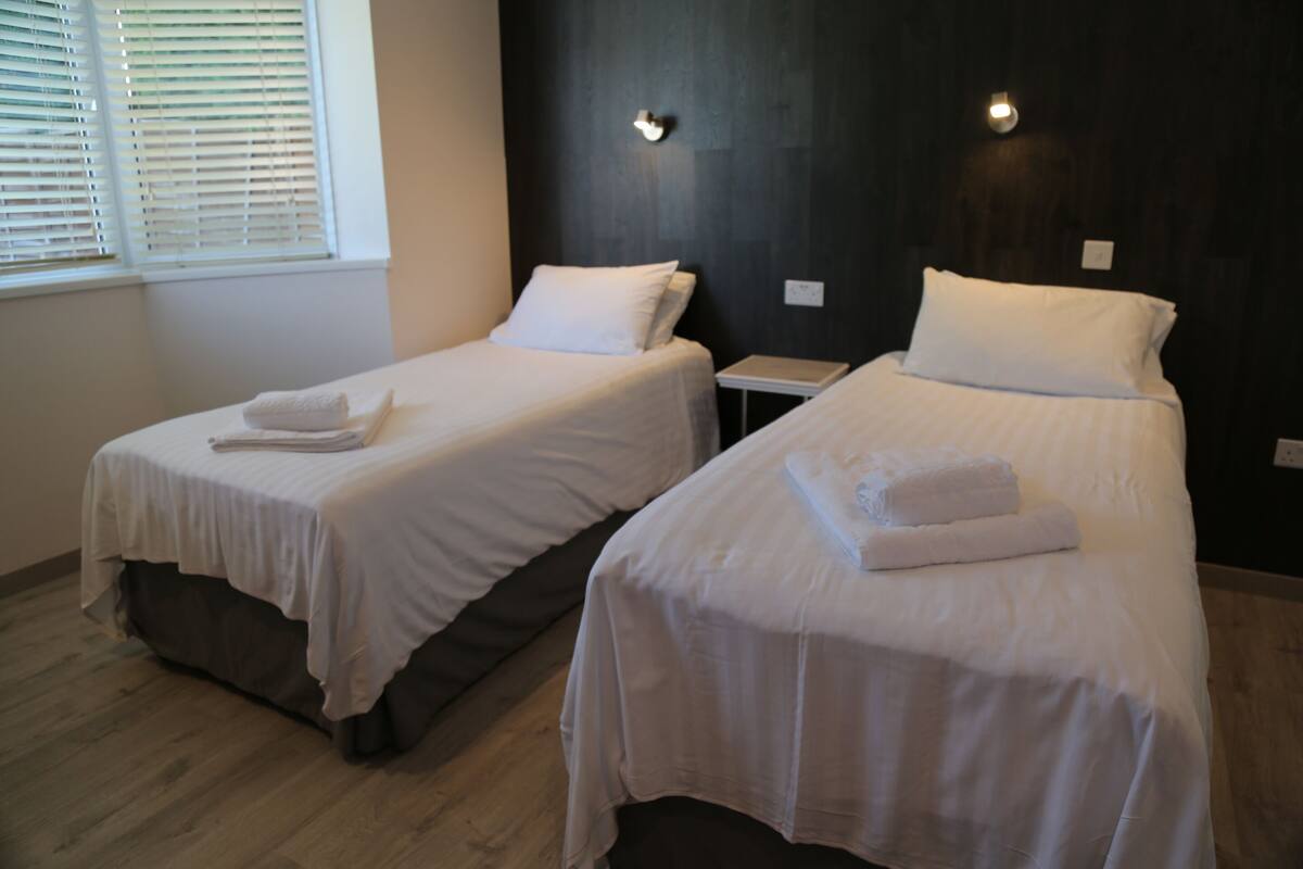 Duas camas de solteiro com roupa de cama e almofadas brancas, uma pequena mesa de cabeceira e um aplique na parede.