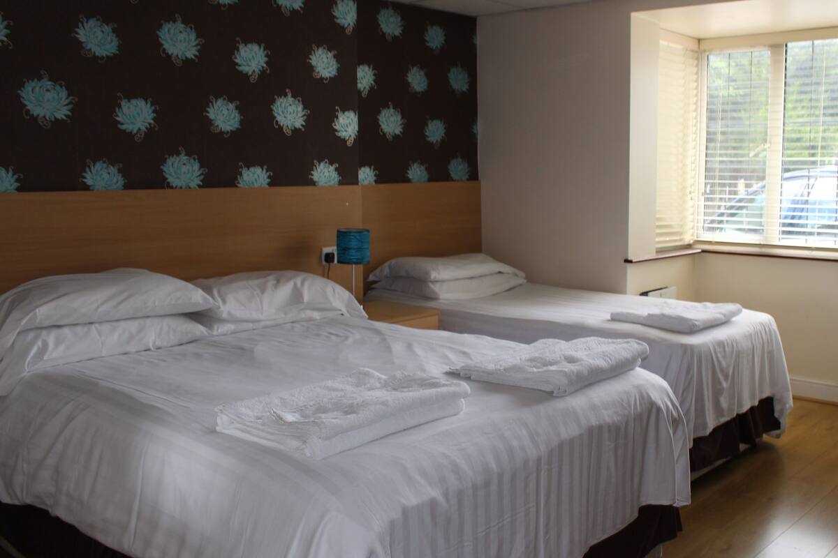 Quarto de hotel com duas camas, uma cabeceira de madeira, parede com padrão floral azul e uma janela com persianas.
