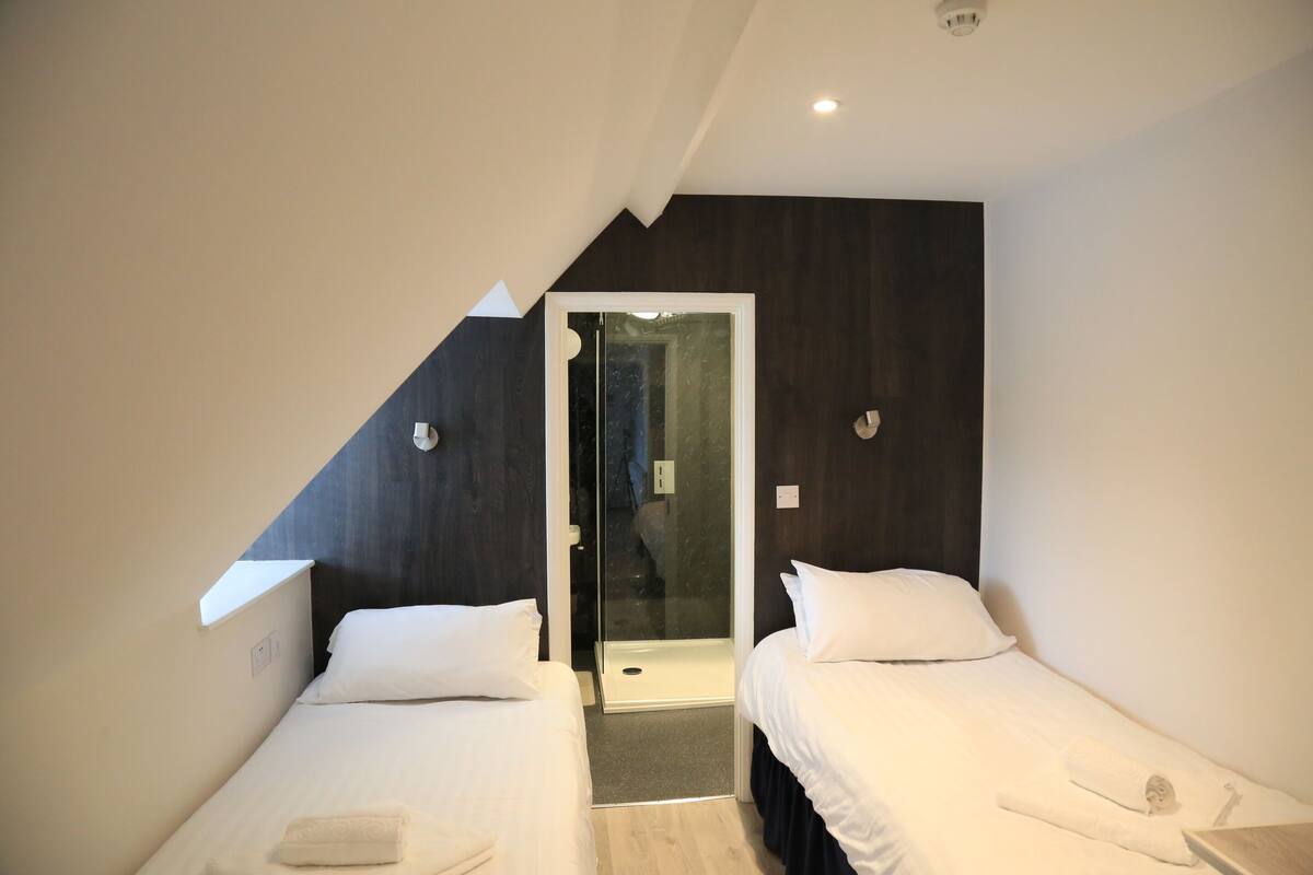 Quarto de hotel moderno com duas camas individuais, box de vidro para o chuveiro e teto inclinado.