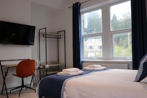 Quarto de hotel com uma cama, uma escrivaninha com cadeira, televisão e janela com cortinas.