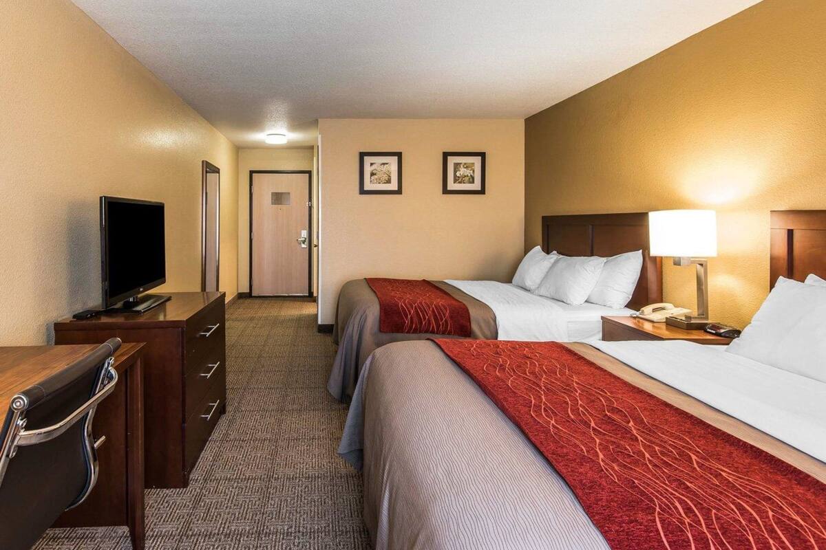 Quarto de hotel com duas camas, uma escrivaninha com televisão, uma porta e um quadro fixado na parede.