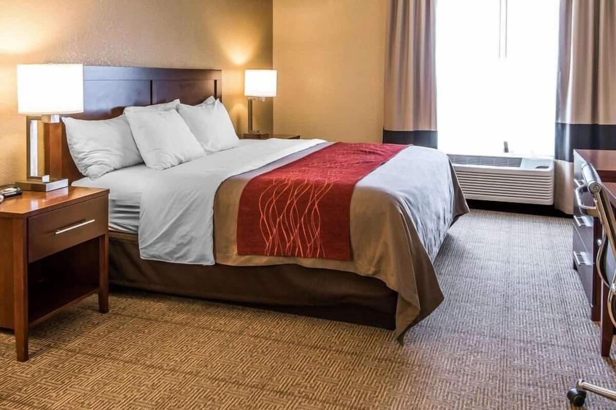 Quarto de hotel com cama, duas luminárias de cabeceira, mesa de cabeceira, escrivaninha, ar condicionado e janela com cortinas.