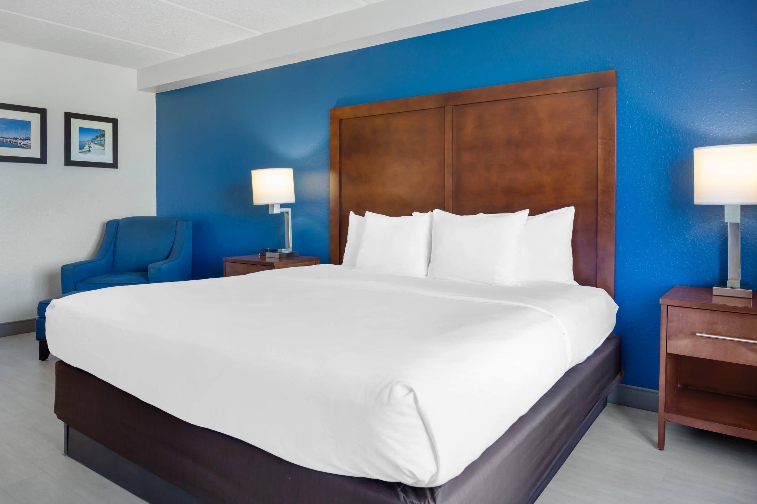 Quarto de hotel com uma cama grande, duas mesas de cabeceira com abajures, uma poltrona azul e quadros nas paredes.