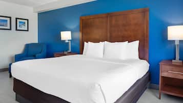 Quarto de hotel com uma cama grande, duas mesas de cabeceira com abajures, uma poltrona azul e quadros nas paredes.