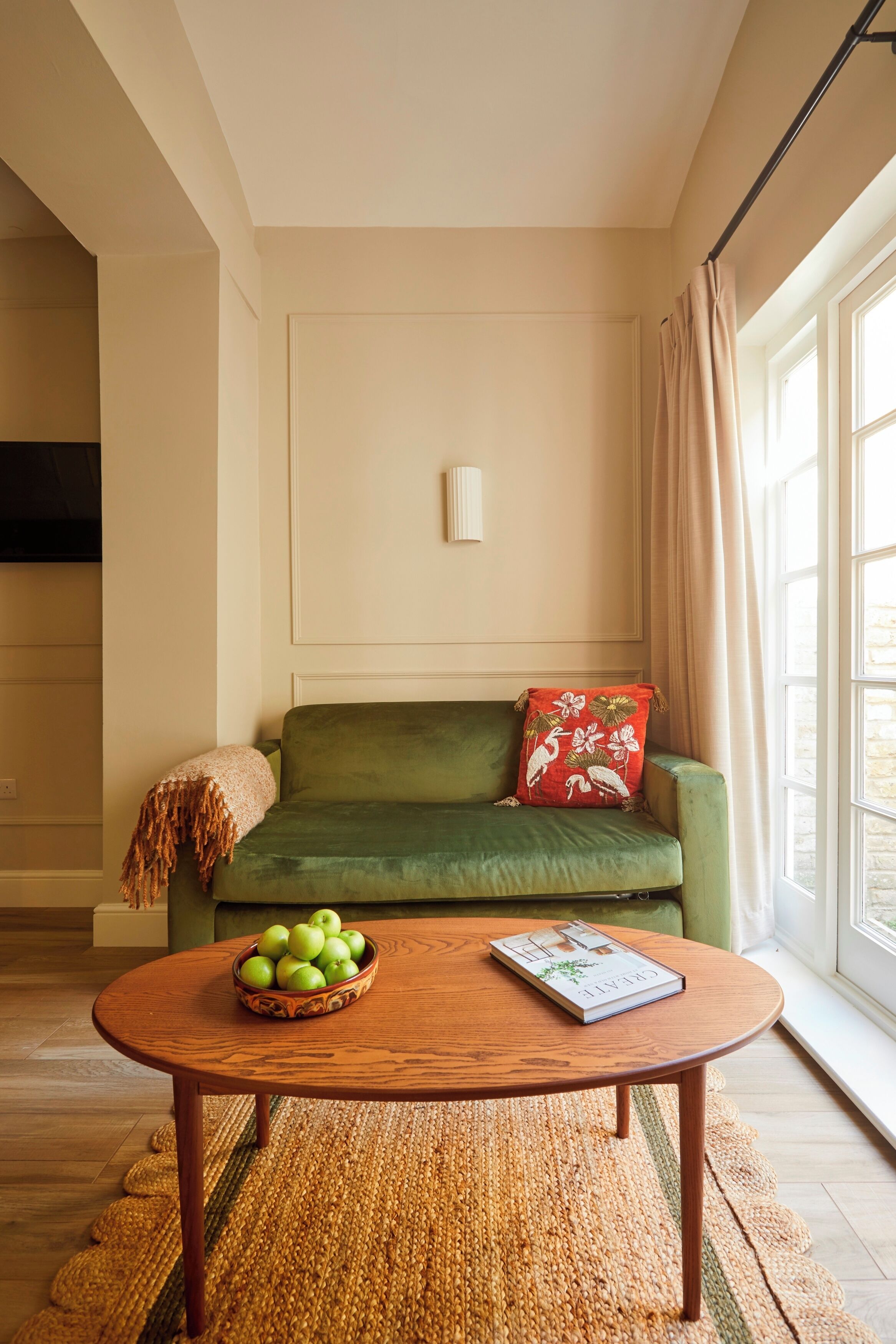 Uma sala de estar acolhedora com um sofá verde, uma mesa de centro de madeira com uma tigela de maçãs e um livro, e uma janela grande com cortinas.
