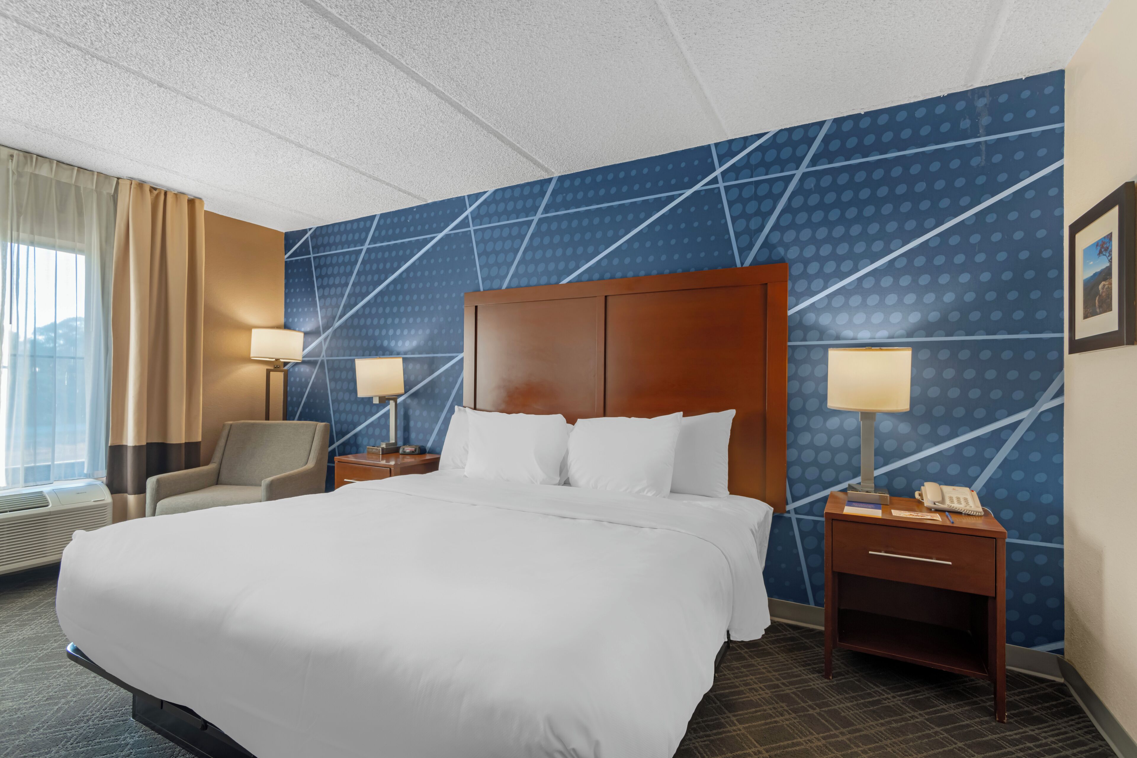 Quarto de hotel com uma cama grande, duas mesas de cabeceira com luminárias, uma cadeira e uma parede com padrão geométrico.