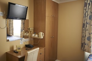 Quarto de hotel com guarda-roupa de madeira, televisão na parede, secretária com telefone, espelho e janela com cortinas florais.