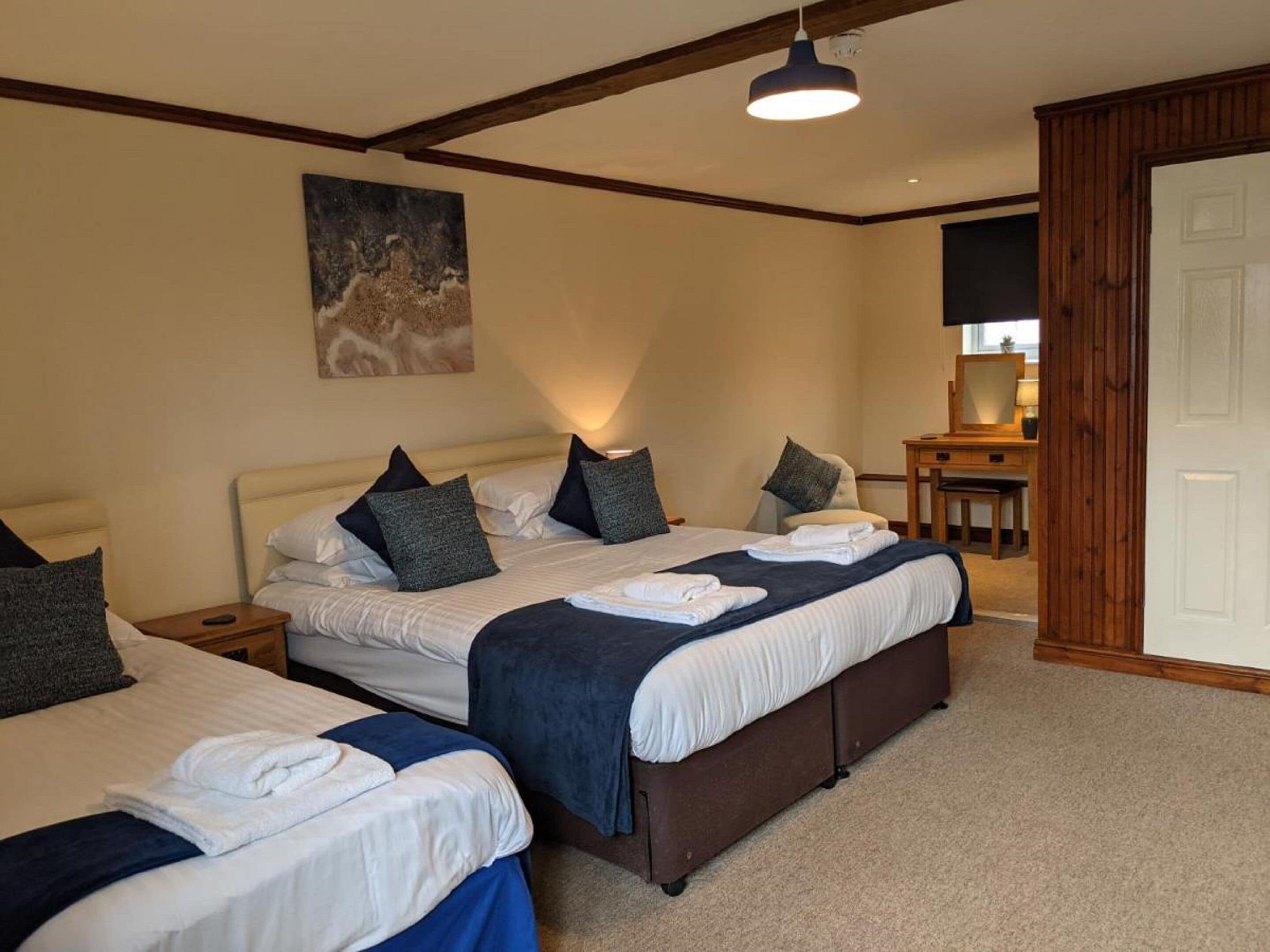 Quarto de hotel com duas camas, um quadro na parede, uma porta de madeira e uma escrivaninha com abajur.