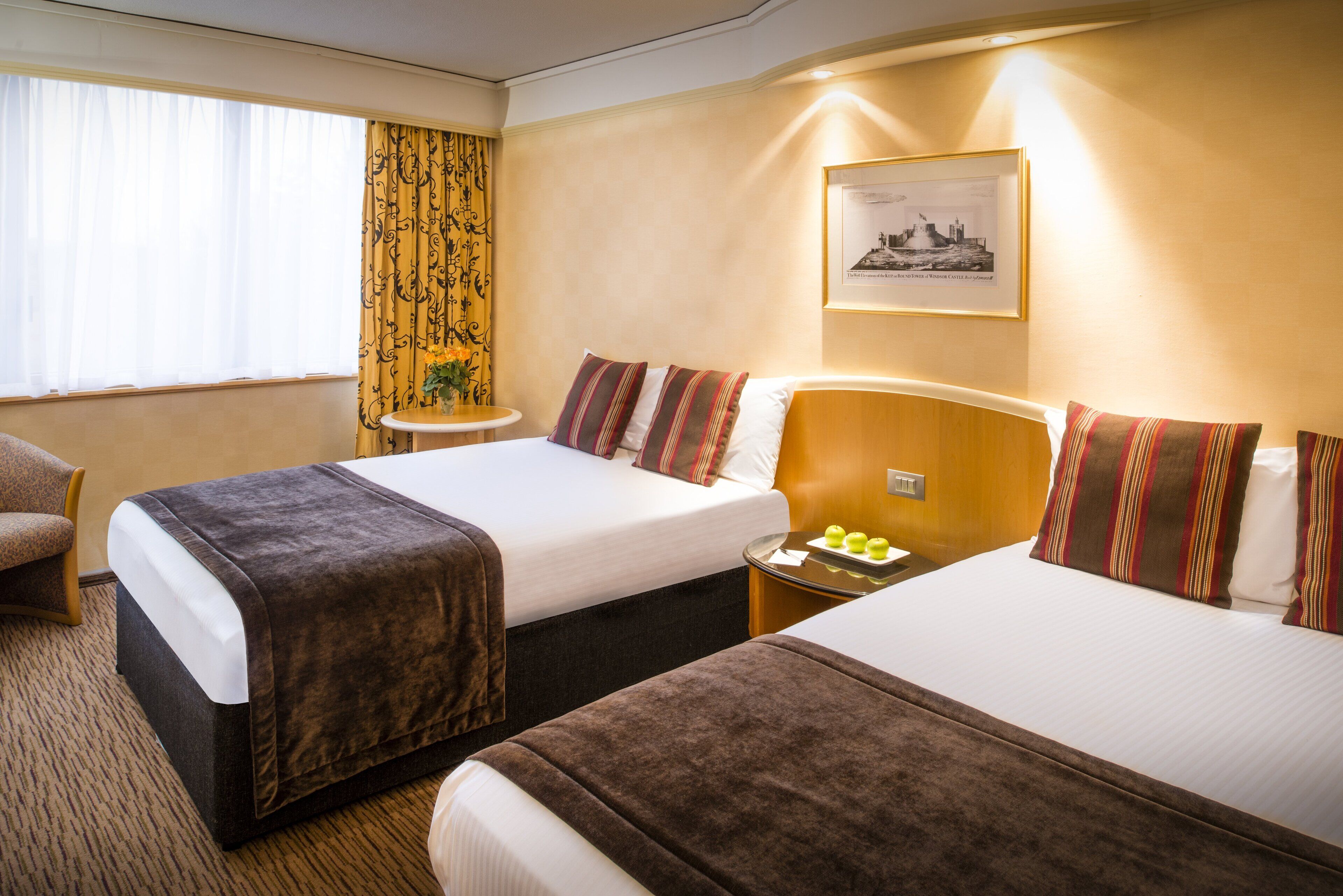 Quarto de hotel com duas camas, uma mesa redonda pequena com um vaso de flores e um quadro na parede.