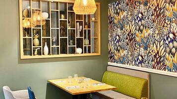 Uma área de refeições com uma mesa de madeira, cadeiras e um banco. Há uma janela grande com padrão quadriculado e um quadro abstrato colorido na parede.