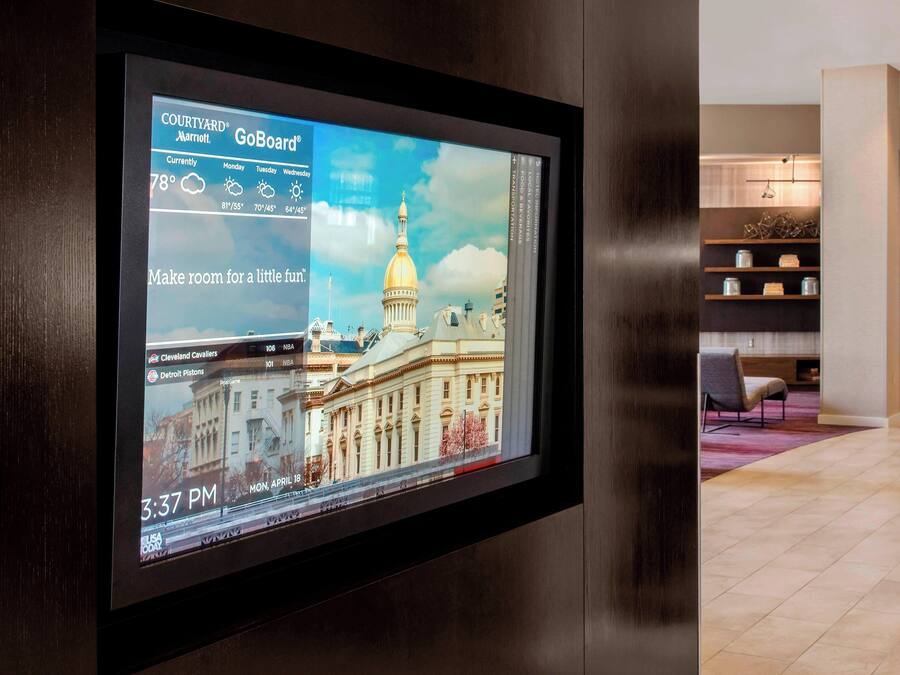 Quarto de hotel com um grande ecrã digital exibindo a previsão do tempo e vista para um edifício histórico.