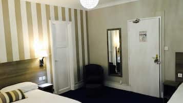 Quarto de hotel com cama, cadeira, escrivaninha, luminária, espelho e porta.