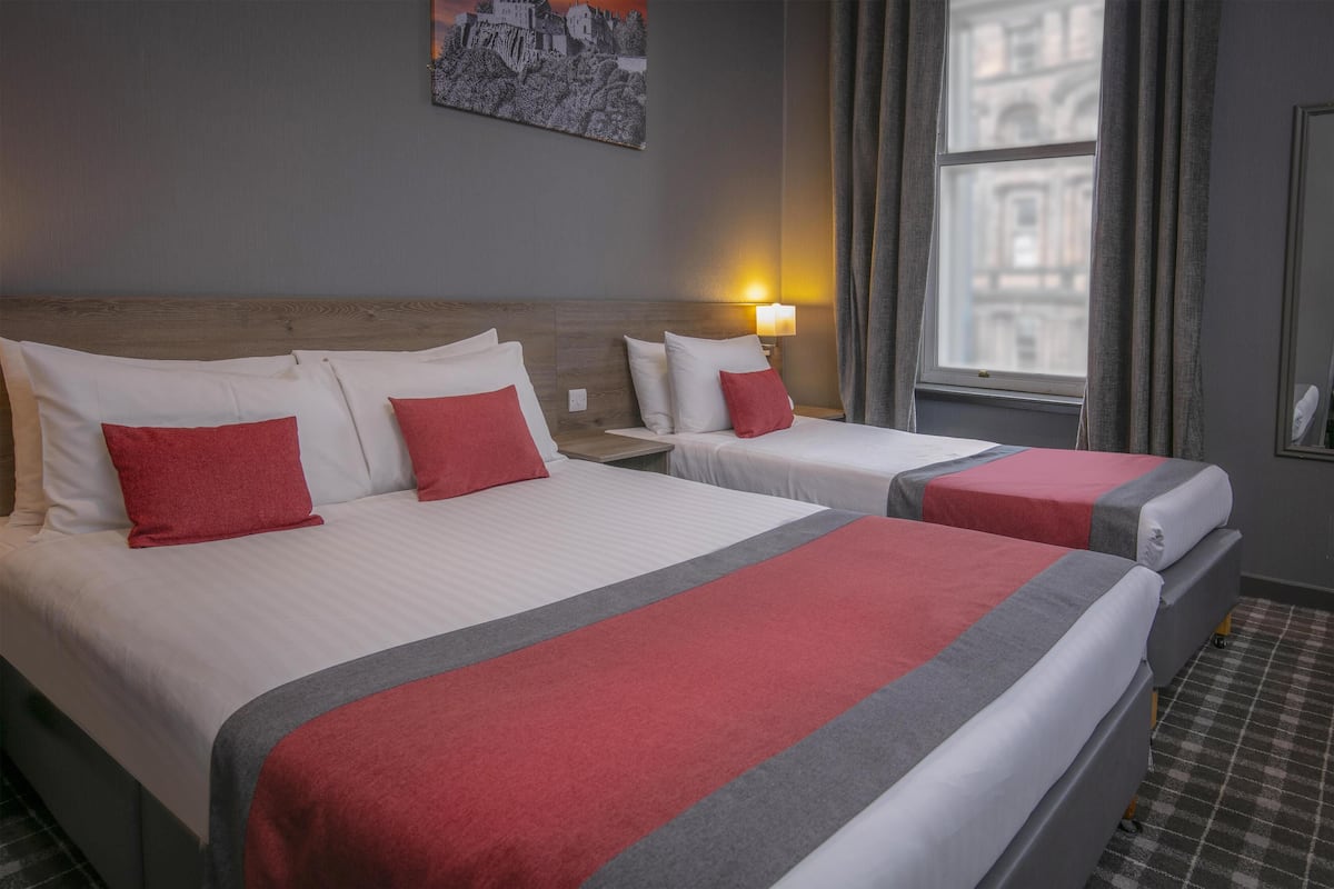 Quarto de hotel com duas camas, uma mesinha de cabeceira, uma luminária, janela com cortinas e um quadro na parede.
