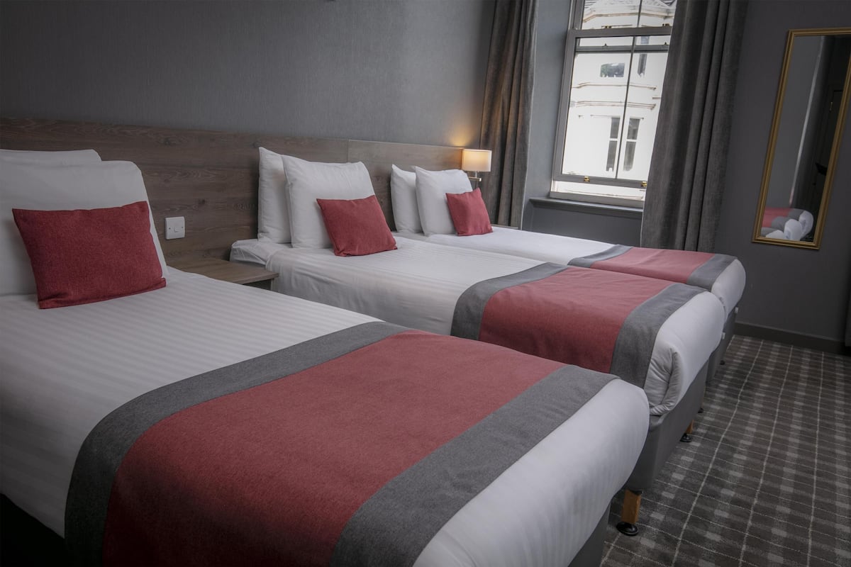 Quarto de hotel com três camas, cada uma com roupa de cama branca e detalhes vermelhos, uma janela com cortinas e piso quadriculado.