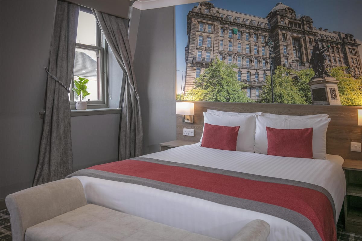 Quarto de hotel com uma cama grande, uma janela com cortinas, uma mesa de cabeceira, um abajur e vista para um edifício com uma estátua.