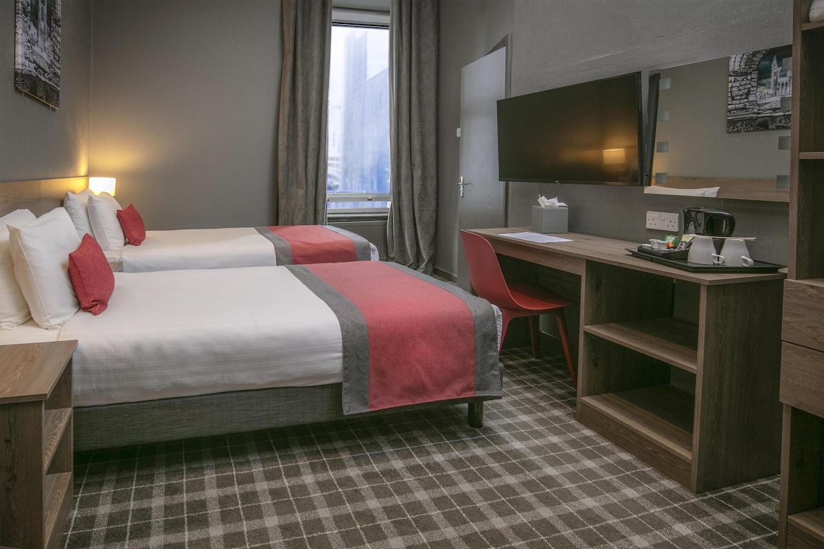 Quarto de hotel com duas camas, uma escrivaninha, uma cadeira, televisão e janela com vista para a cidade.