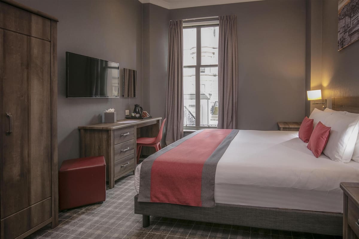 Quarto de hotel com cama, escrivaninha, cadeira, guarda-roupa, televisão, janela com cortinas e mesa de cabeceira com abajur.