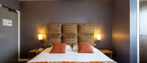 Quarto de hotel com uma cama grande, cabeceira com padrão trançado, duas mesas de cabeceira com luminárias e um edredom acolchoado.