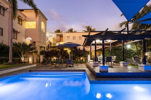 Piscina com espreguiçadeiras e guarda-sóis, cercada por um jardim ajardinado e um edifício com varandas.