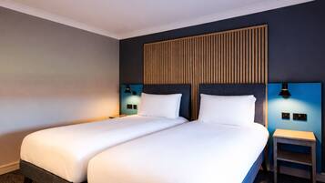 Quarto de hotel com duas camas, painel de cabeceira de madeira, abajures e uma pequena mesa.