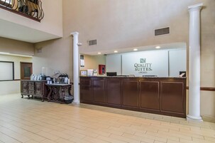 Um balcão de recepção com uma placa que diz 'Quality Suites'. Há um armário de madeira com diversos objetos e uma coluna branca visível à direita.