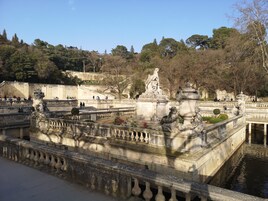 Uma ponte de pedra histórica com estátuas ornamentadas e um espelho d'água, rodeada por árvores e um muro de pedra.