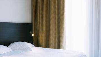 Uma cama bem arrumada com lençóis brancos, cabeceira escura e uma janela grande com cortinas leves.