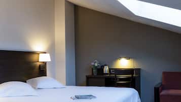 Quarto de hotel com uma cama, duas luminárias laterais, uma mesa de cabeceira com luminária, uma cadeira e um claraboia.