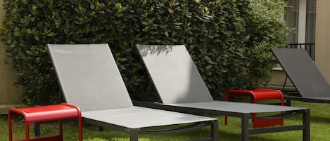 Duas cadeiras de exterior com estrutura de metal e almofadas cinzentas, localizadas numa área relvada com um sequeiro verde denso ao fundo.