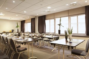Uma sala de conferências com várias mesas e cadeiras, janelas amplas com cortinas e plantas em vasos.