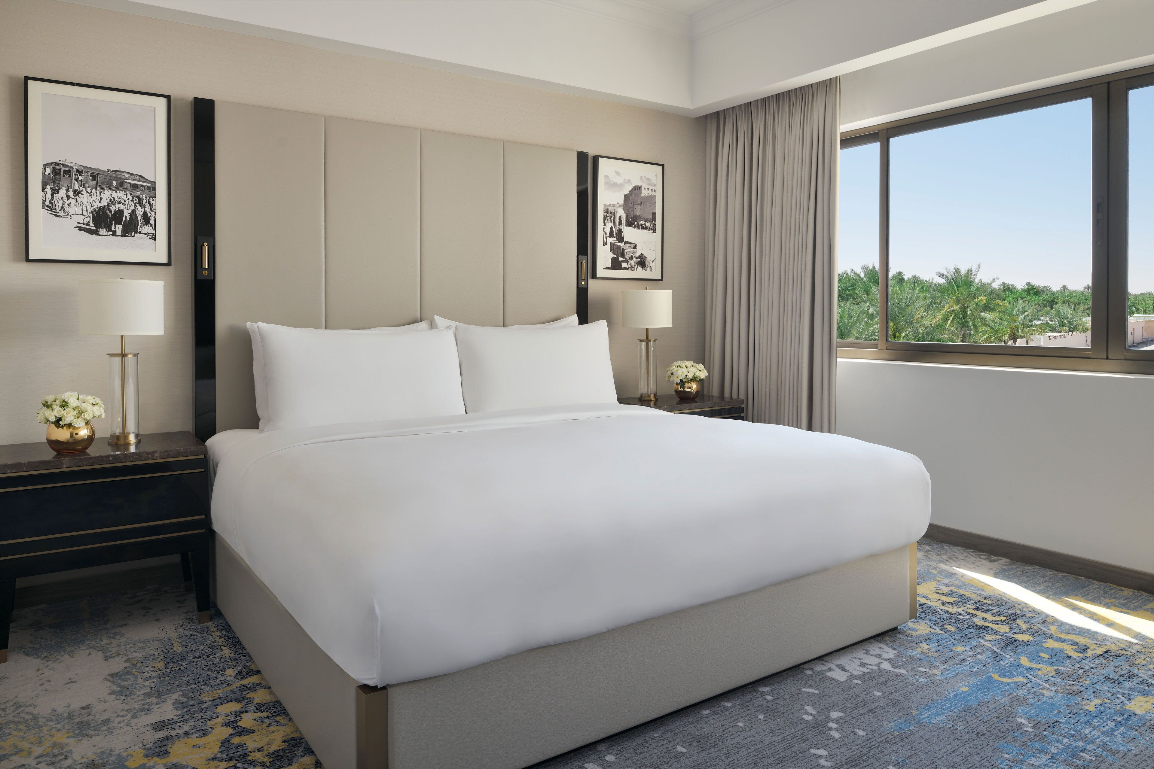Quarto de hotel com uma cama grande, duas mesinhas de cabeceira, uma janela com vista para palmeiras e duas quadros pendurados na parede.