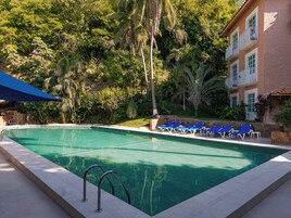 Piscina com águas cristalinas azuis, rodeada por vegetação exuberante e um edifício com varandas.