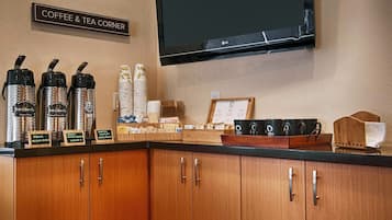 Um espaço com estação de café e chá, incluindo diversos distribuidores de café, uma placa indicando o cantinho do café e chá, e uma televisão fixada na parede.