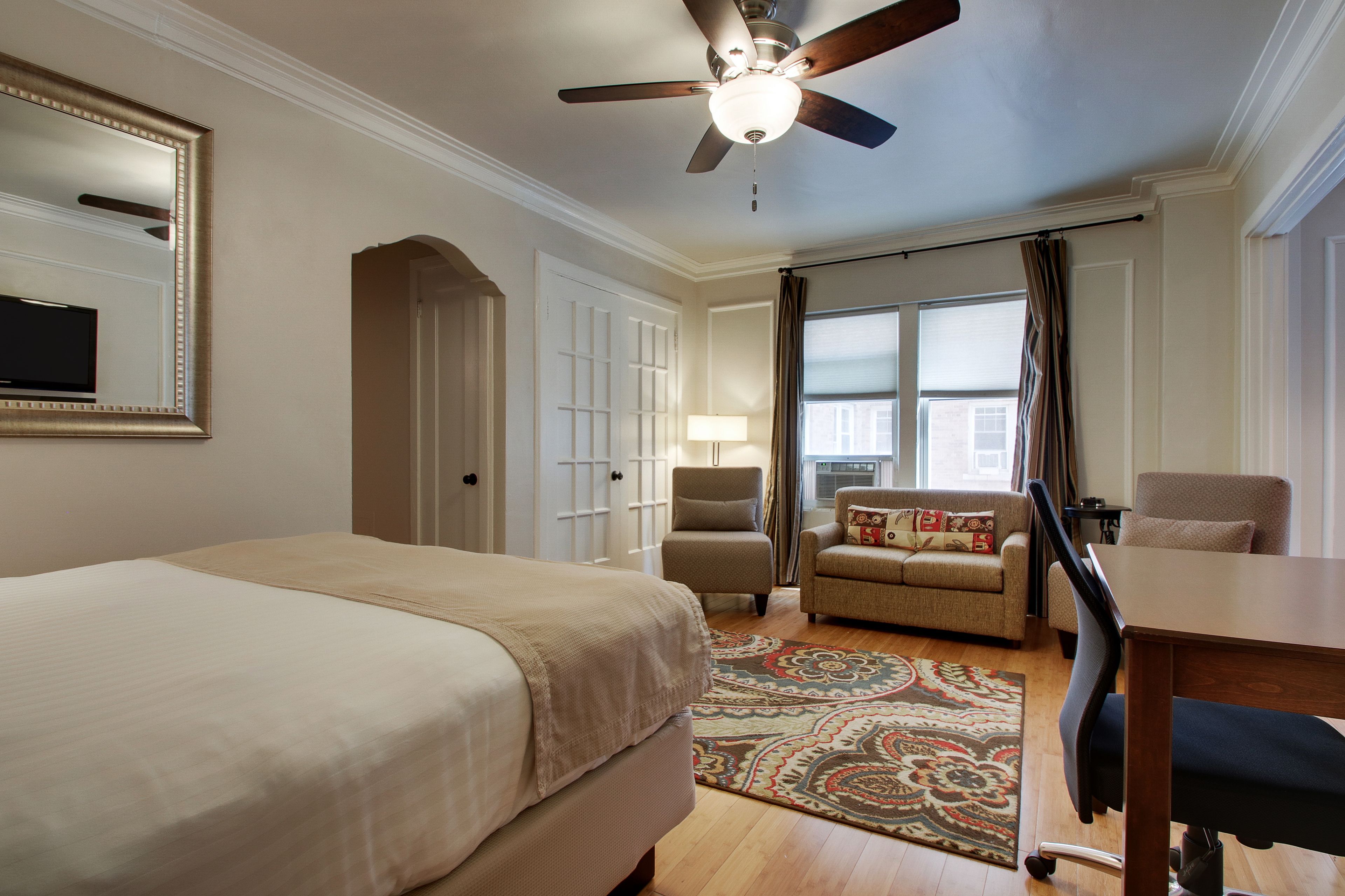 Quarto de hotel com cama, escrivaninha, cadeira, sofá, ventoinha no teto e uma janela grande com cortinas.