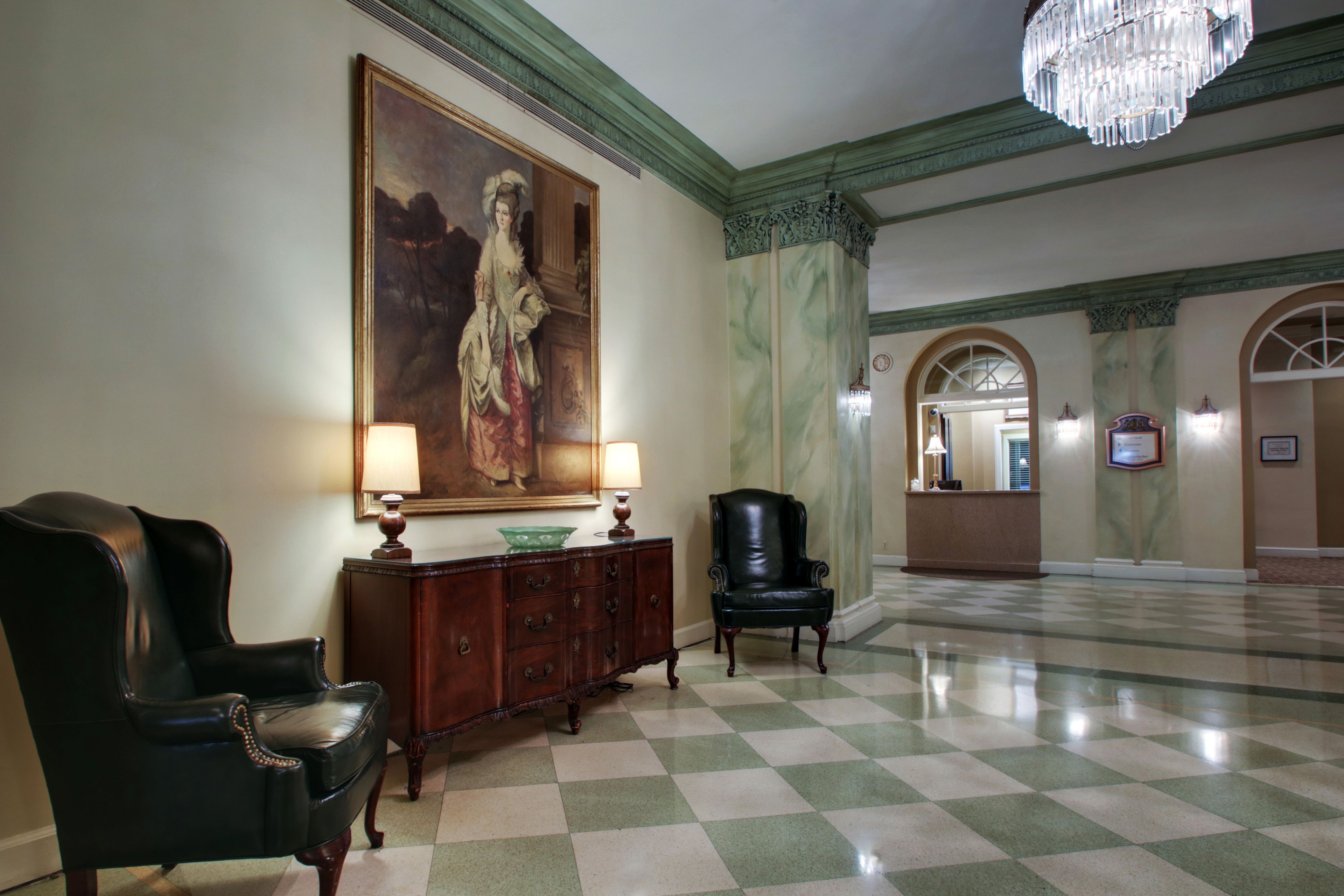 Um hall com piso quadriculado, uma pintura grande, um lustre e um balcão de recepção.
