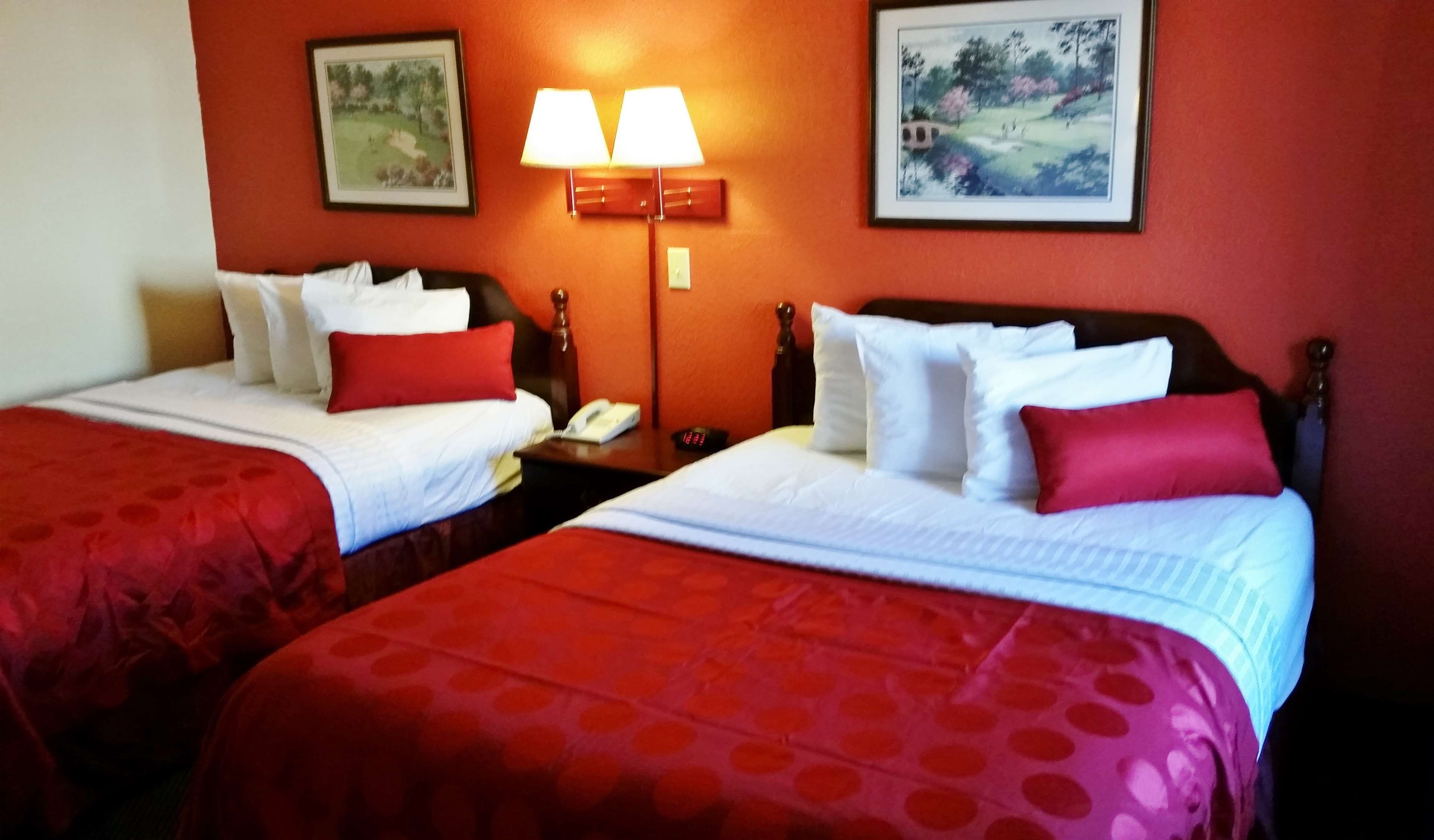 Quarto de hotel com duas camas, roupa de cama vermelha, mesa de cabeceira com abajur e duas quadros na parede.
