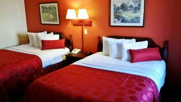 Quarto de hotel com duas camas, roupa de cama vermelha, mesa de cabeceira com abajur e duas quadros na parede.