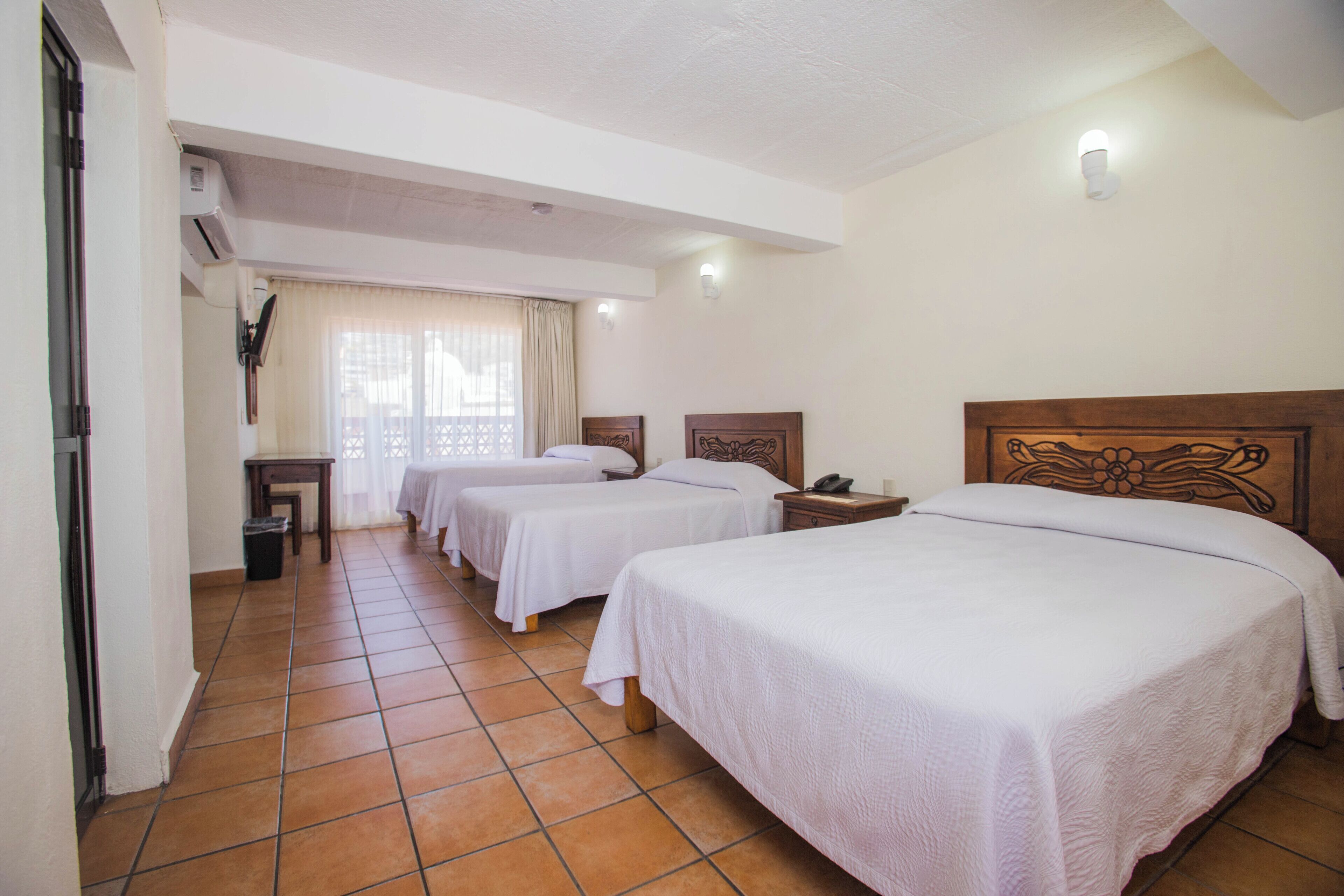 Quarto de hotel com três camas, uma mesinha e uma janela com cortinas.