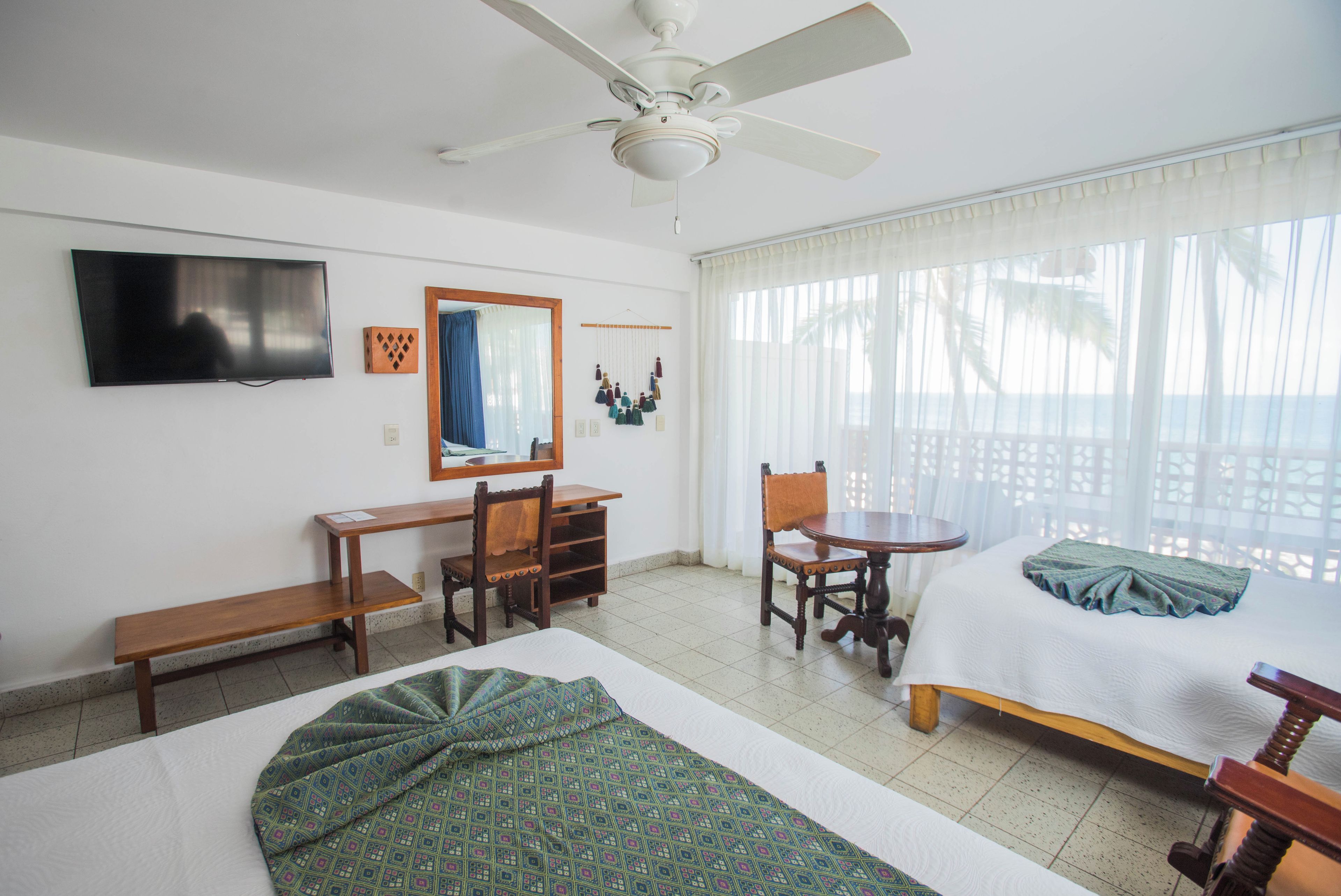 Quarto de hotel com duas camas, uma escrivaninha, uma cadeira, uma televisão e um ventilador de teto.