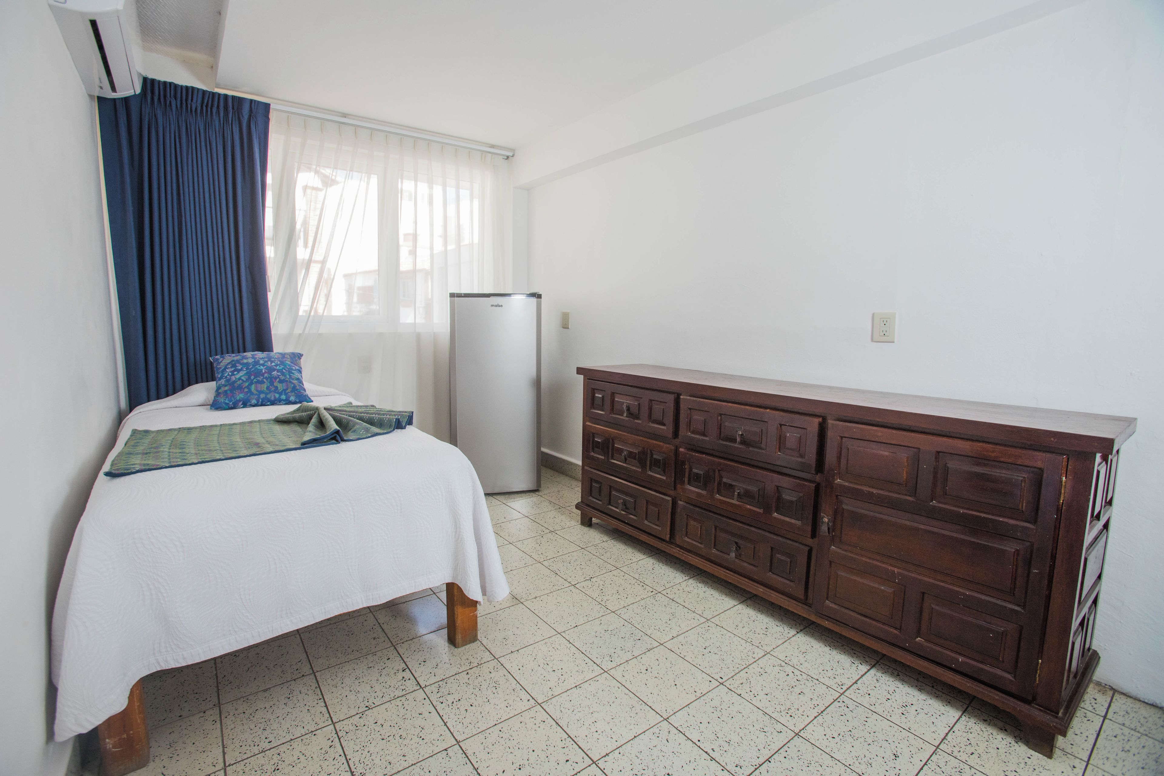 Quarto com cama, cômoda, janela com cortinas e geladeira.