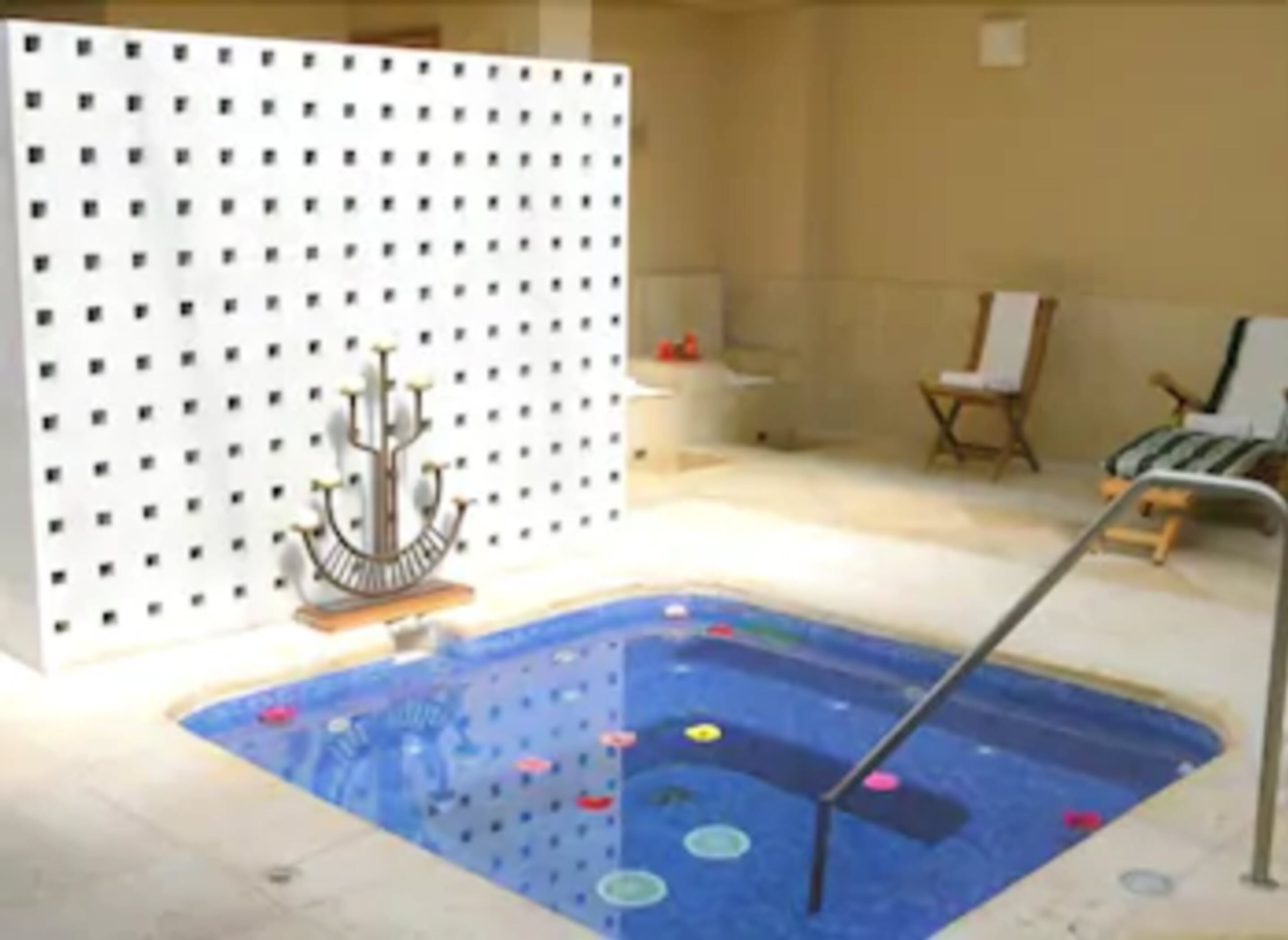 Pequena piscina interior com escada, uma fonte decorativa e parede branca com buracos.