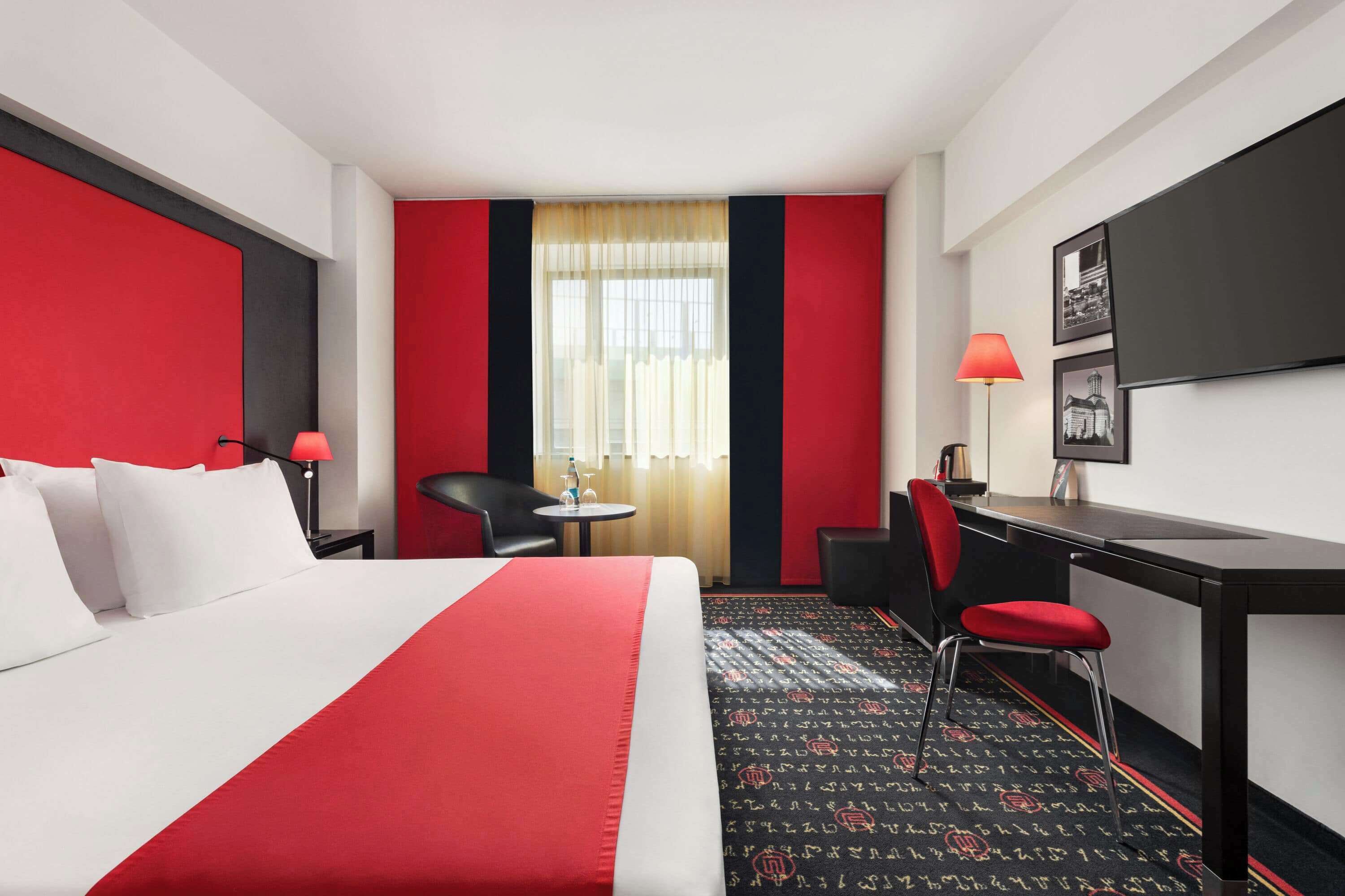 Quarto de hotel com uma cama grande, uma escrivaninha com cadeira, uma mesinha com cadeira e uma televisão fixada na parede.