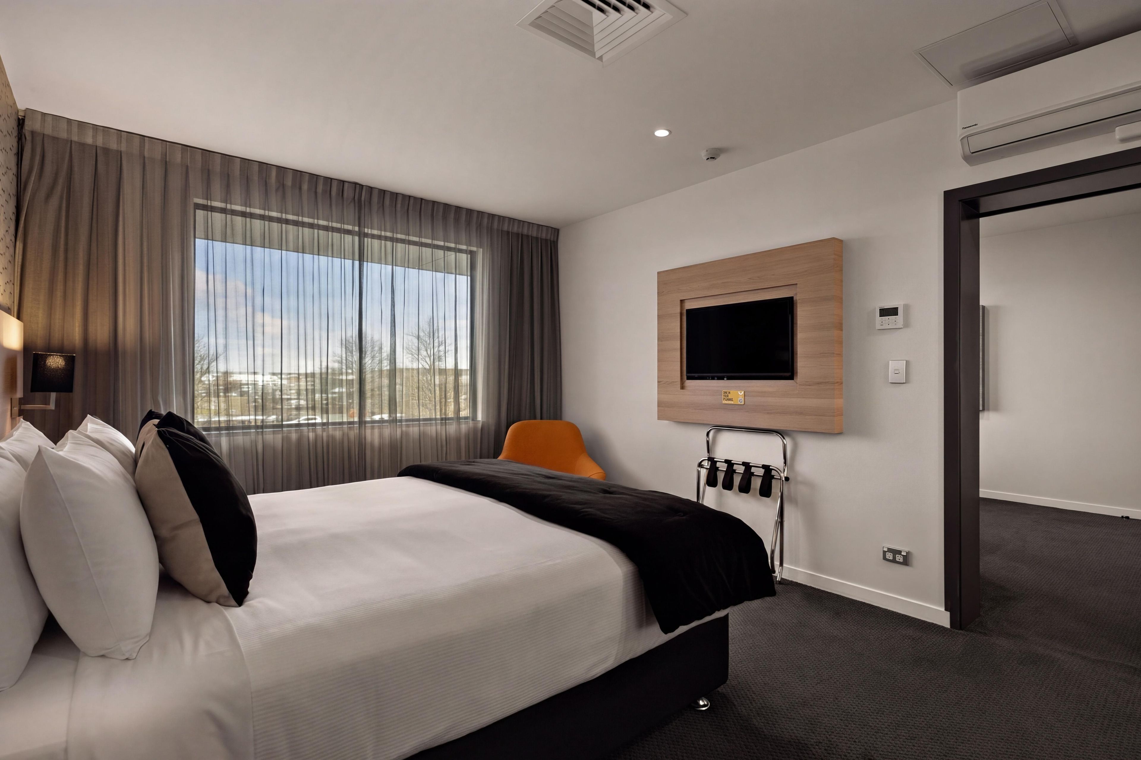 Quarto de hotel com cama, televisão na parede, uma cadeira e vista para o exterior pela janela.