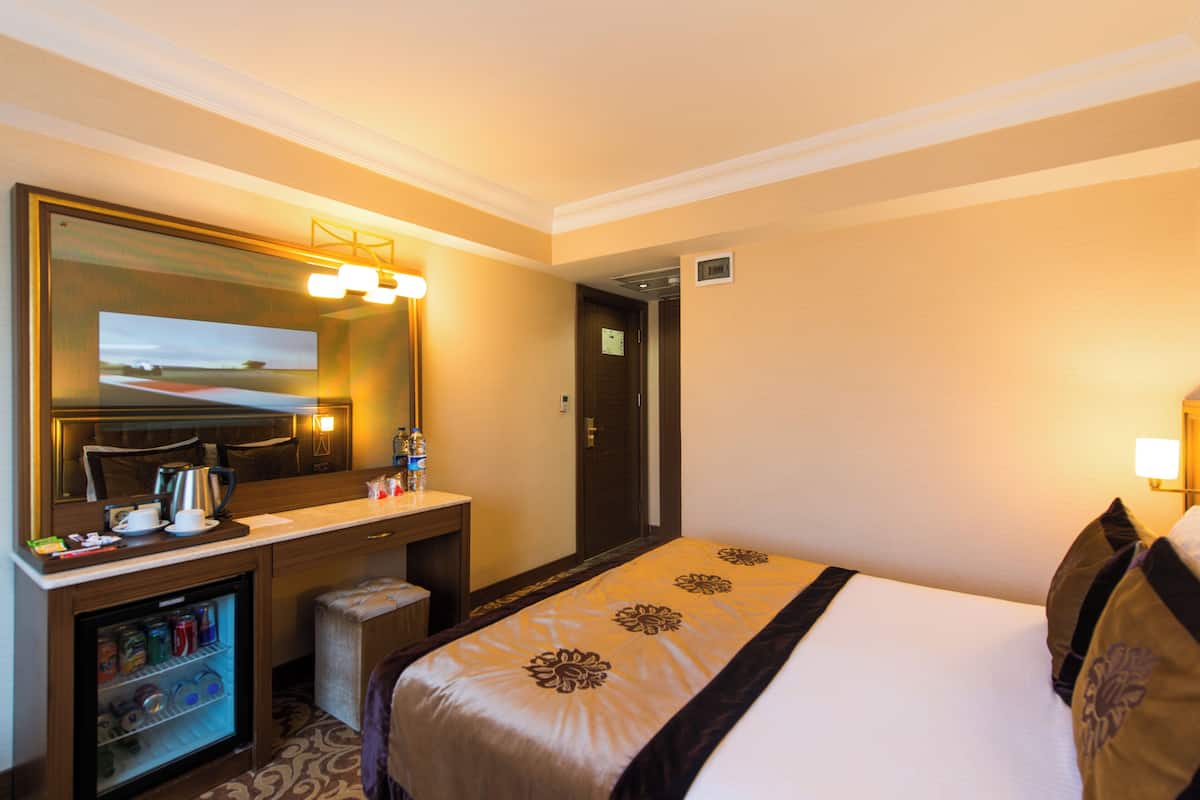 Quarto de hotel com uma cama, uma escrivaninha com espelho, um minibar e uma porta que dá para outro quarto.